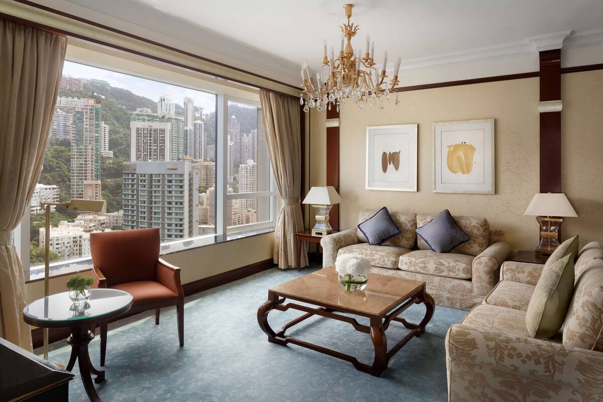 Quarto de hotel com uma grande janela que oferece vista da cidade, um candelabro, um conjunto de sofás e uma pequena mesa com um vaso.