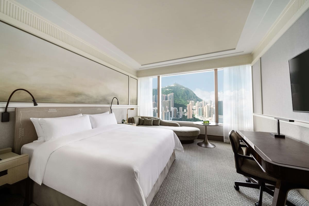 Quarto de hotel com uma cama grande, uma escrivaninha e vista para a paisagem urbana através de amplas janelas.
