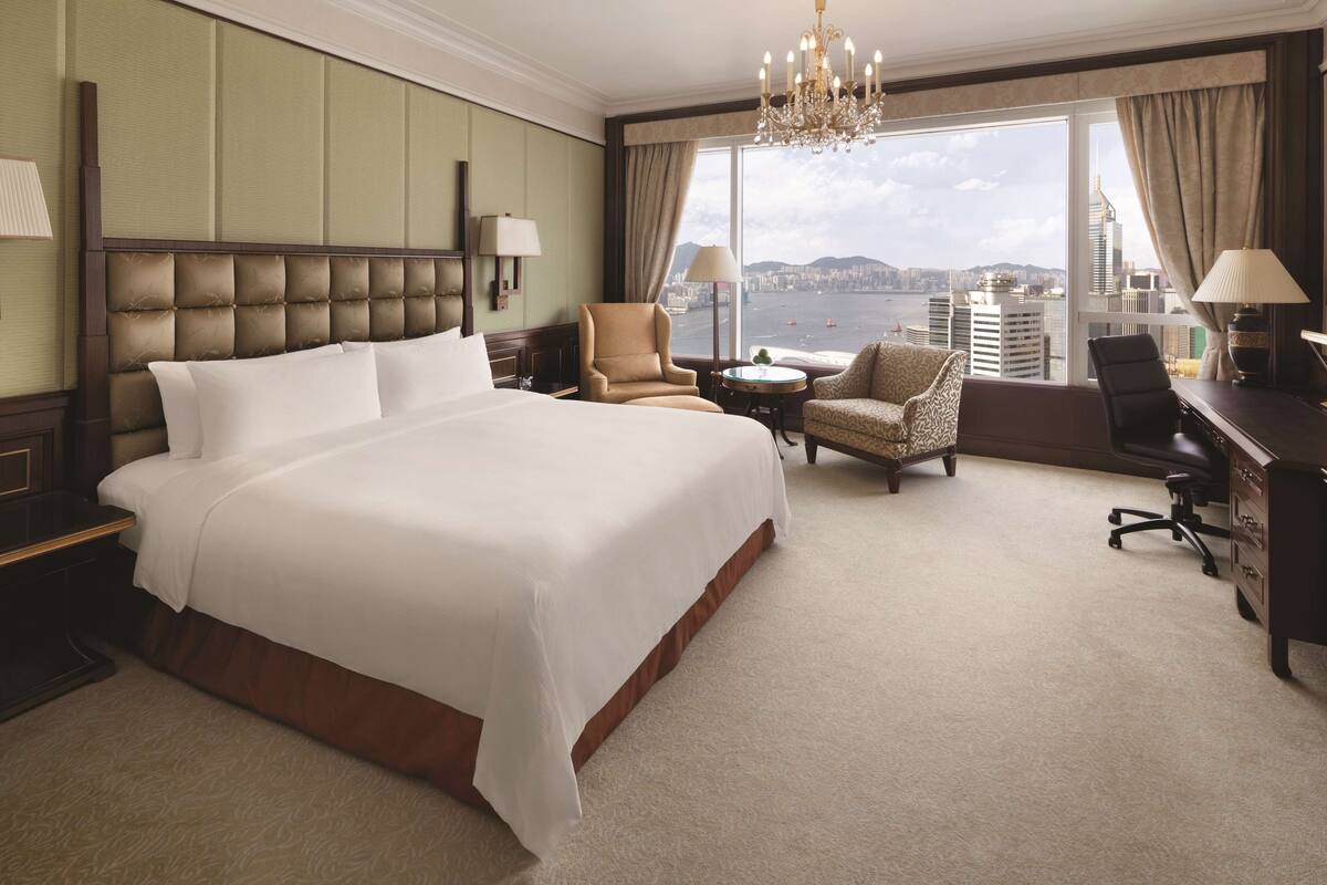 Quarto de hotel com uma cama grande, duas poltronas, uma escrivaninha e vista para a cidade.