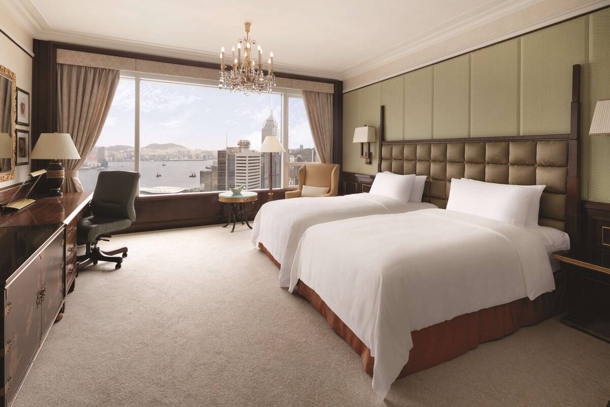 Quarto de hotel com duas camas, uma escrivaninha com candeeiro, uma cadeira, uma mesinha e vista para a paisagem urbana.