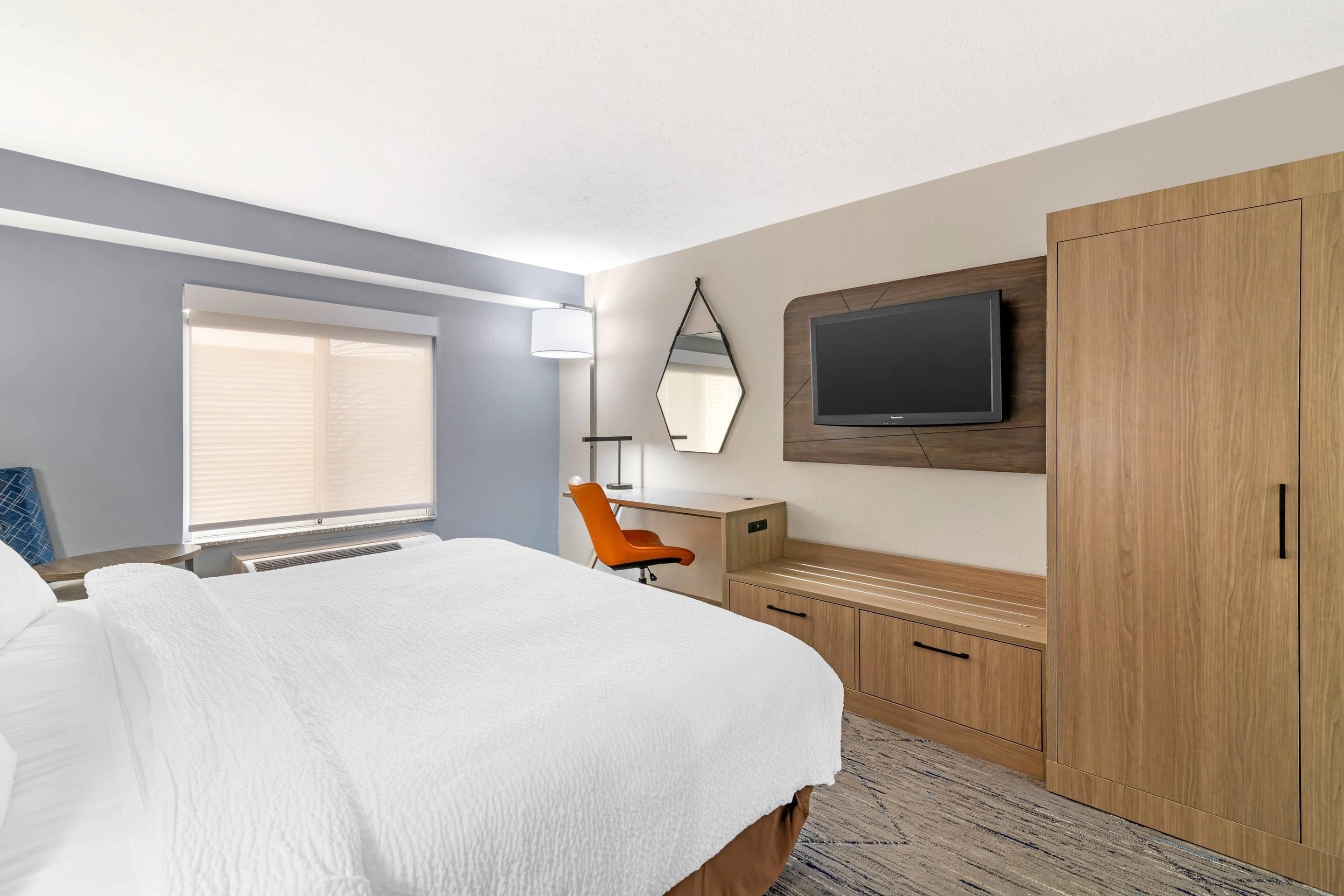 Quarto de hotel com uma cama grande, uma secretária com cadeira, uma televisão na parede e um guarda-roupa.