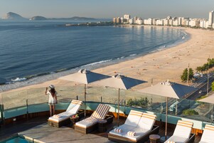 Área de piscina no terraço com espreguiçadeiras e guarda-sóis, com vista para uma praia.