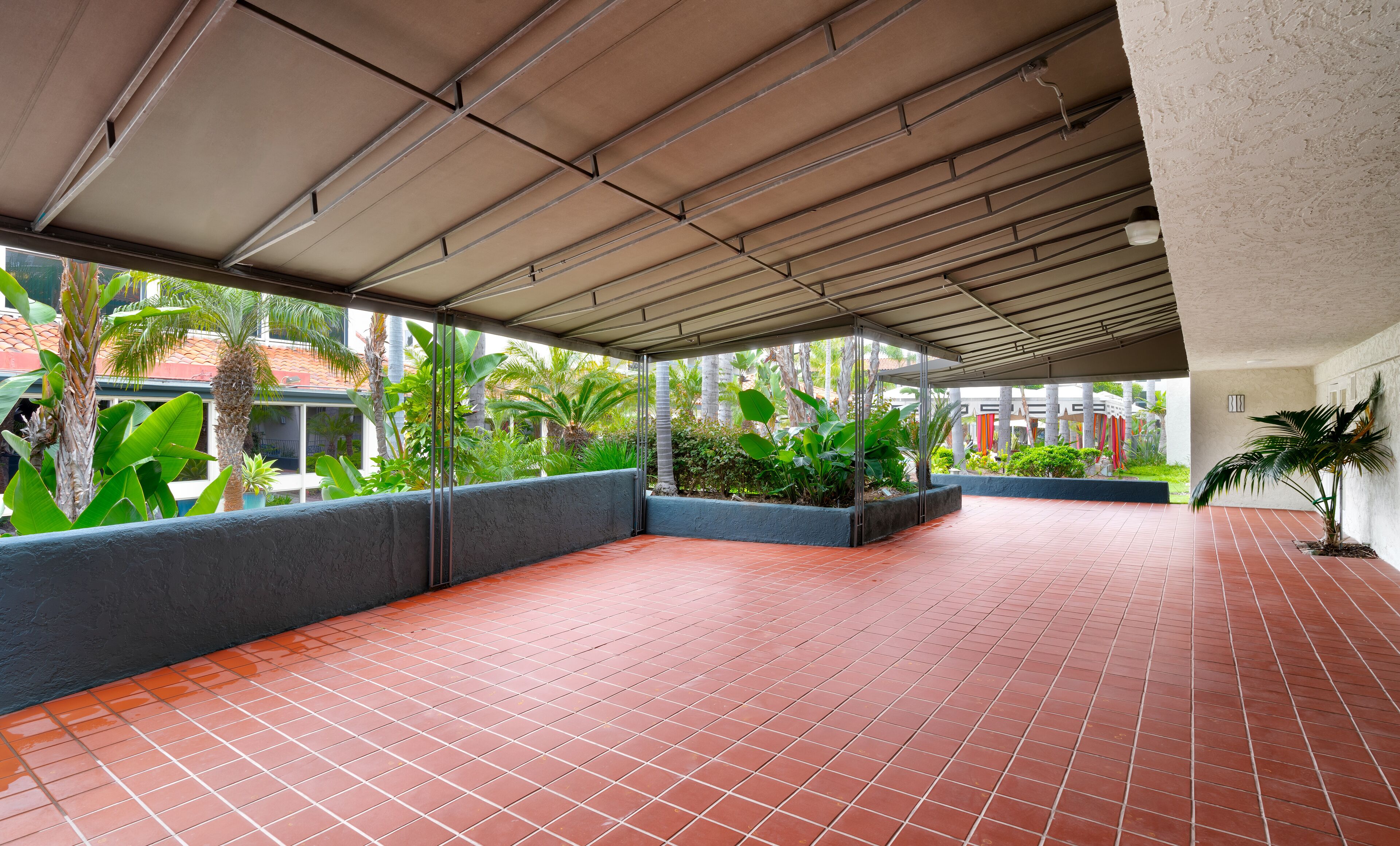 Uma área externa coberta com piso de azulejos, vasos com plantas tropicais e vista para edifícios ao fundo.