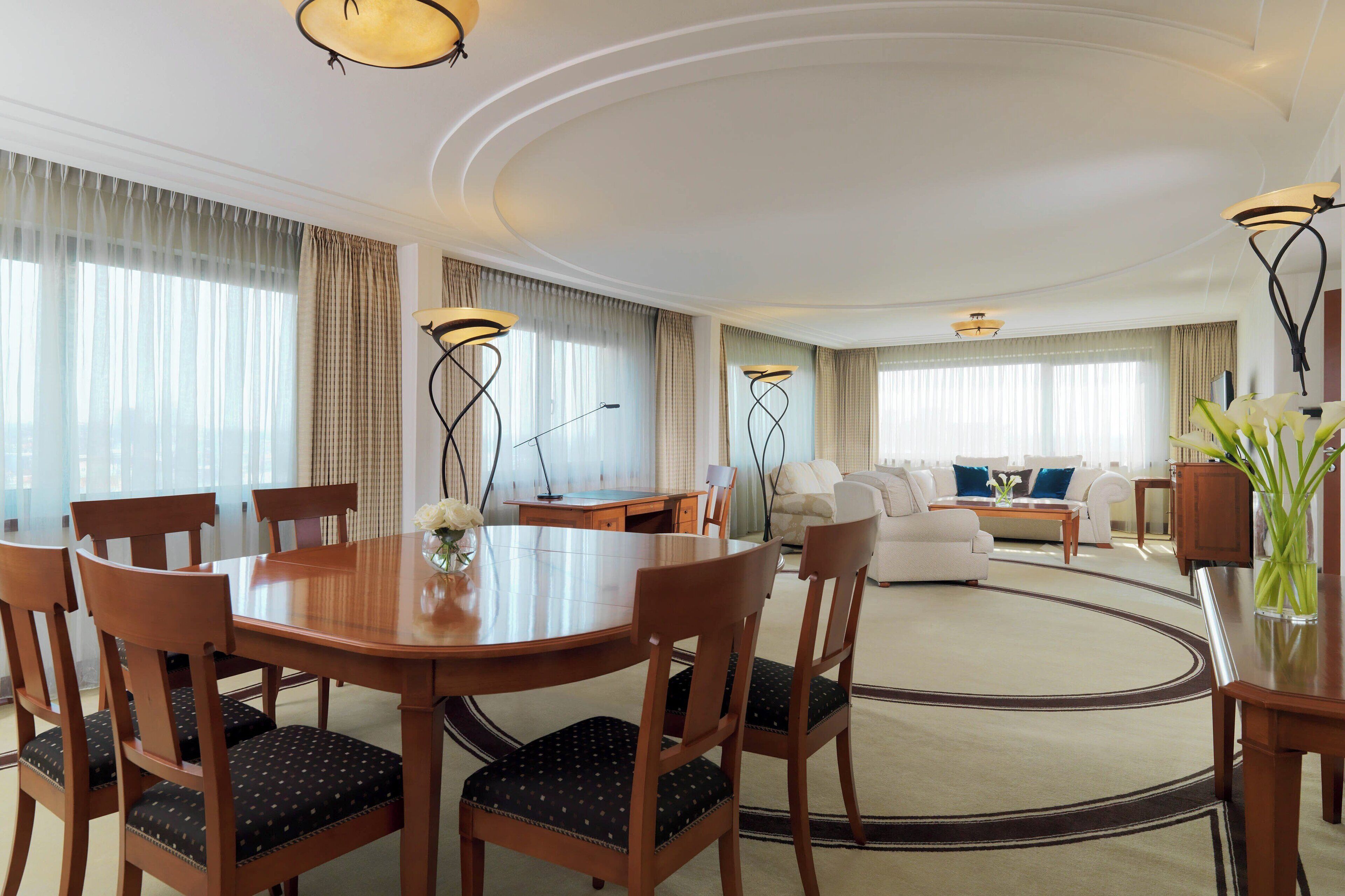 Quarto de hotel com área de jantar, um conjunto de sofás e uma janela grande com cortinas.