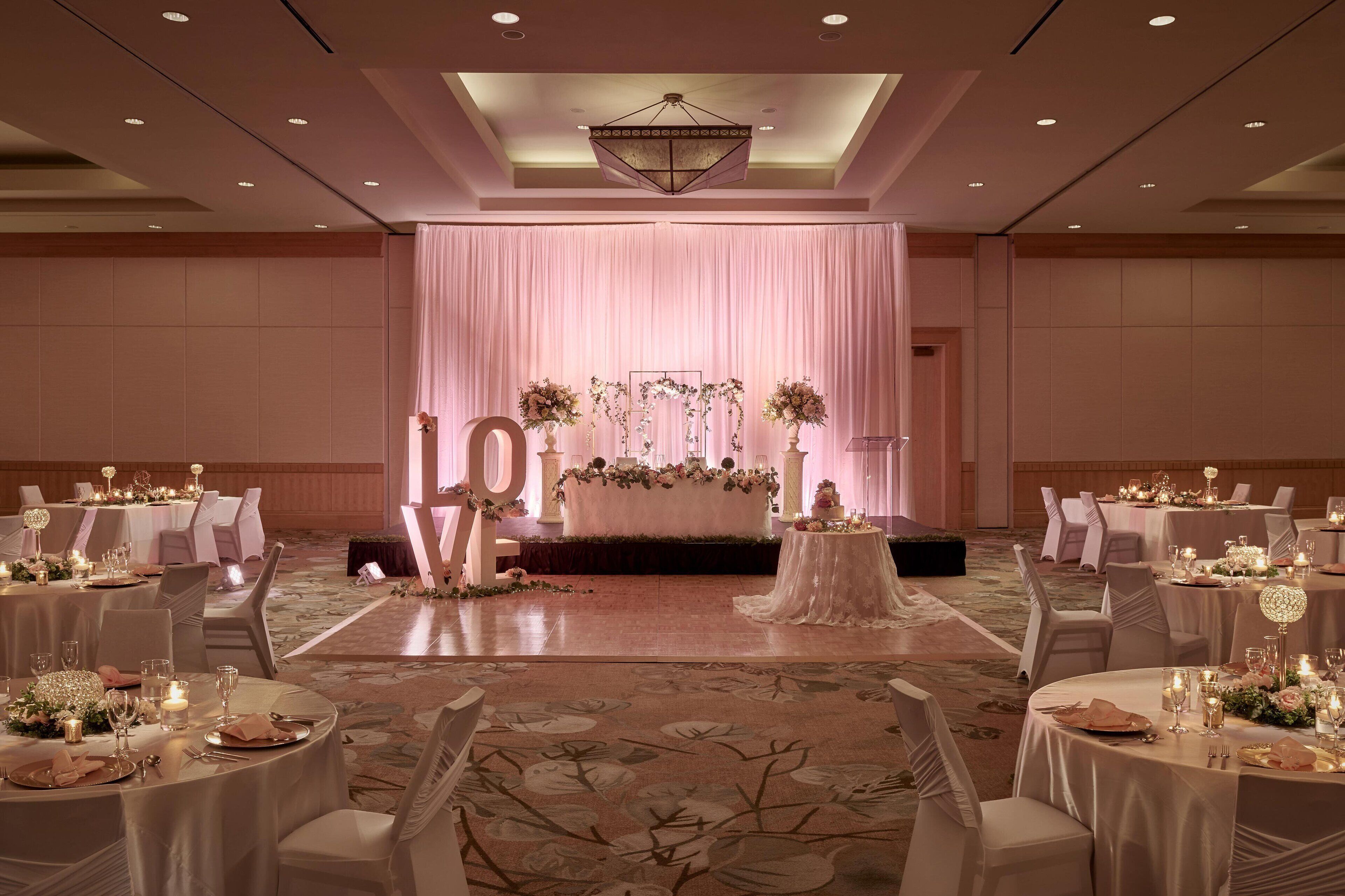 Espaço de recepção de casamento com mesas redondas, cadeiras brancas, palco com fundo rosa, decorações florais e um lustre.