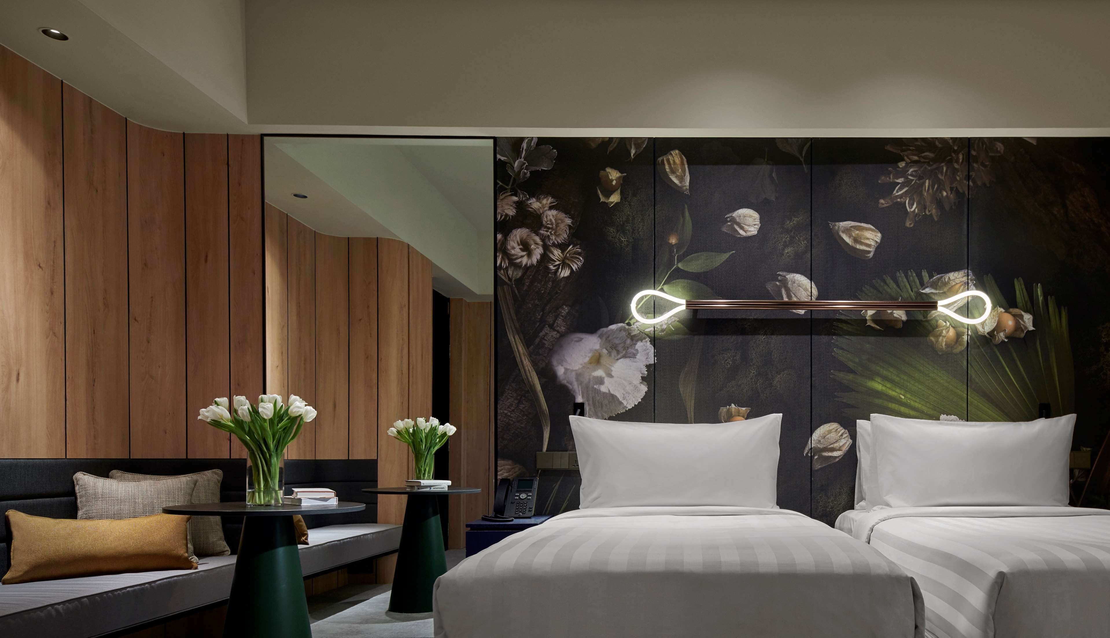 Quarto de hotel moderno com parede de painel de madeira, um espelho grande, uma cama com roupa de cama branca, uma mesa de cabeceira com telefone e um espaço de estar com almofadas e um vaso de flores.