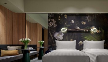 Quarto de hotel moderno com parede de painel de madeira, um espelho grande, uma cama com roupa de cama branca, uma mesa de cabeceira com telefone e um espaço de estar com almofadas e um vaso de flores.