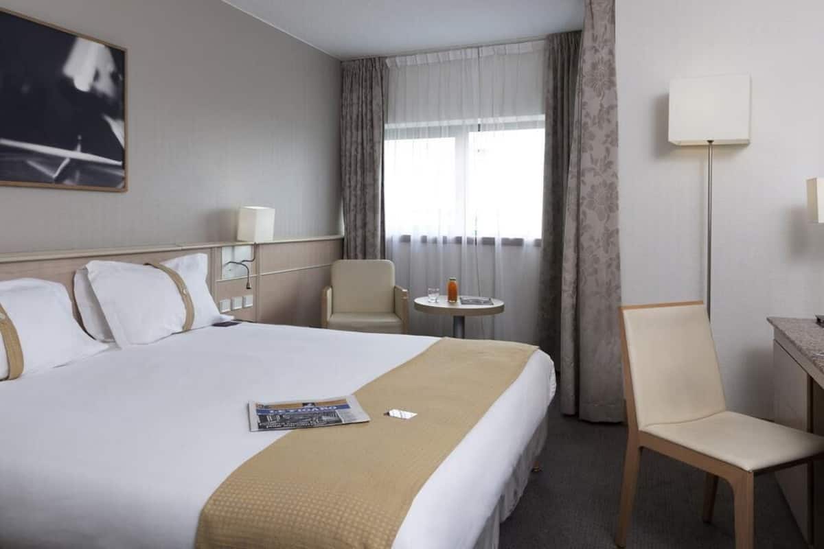 Quarto de hotel com uma cama grande, uma cadeira, uma mesinha, um abajur e uma janela com cortinas.