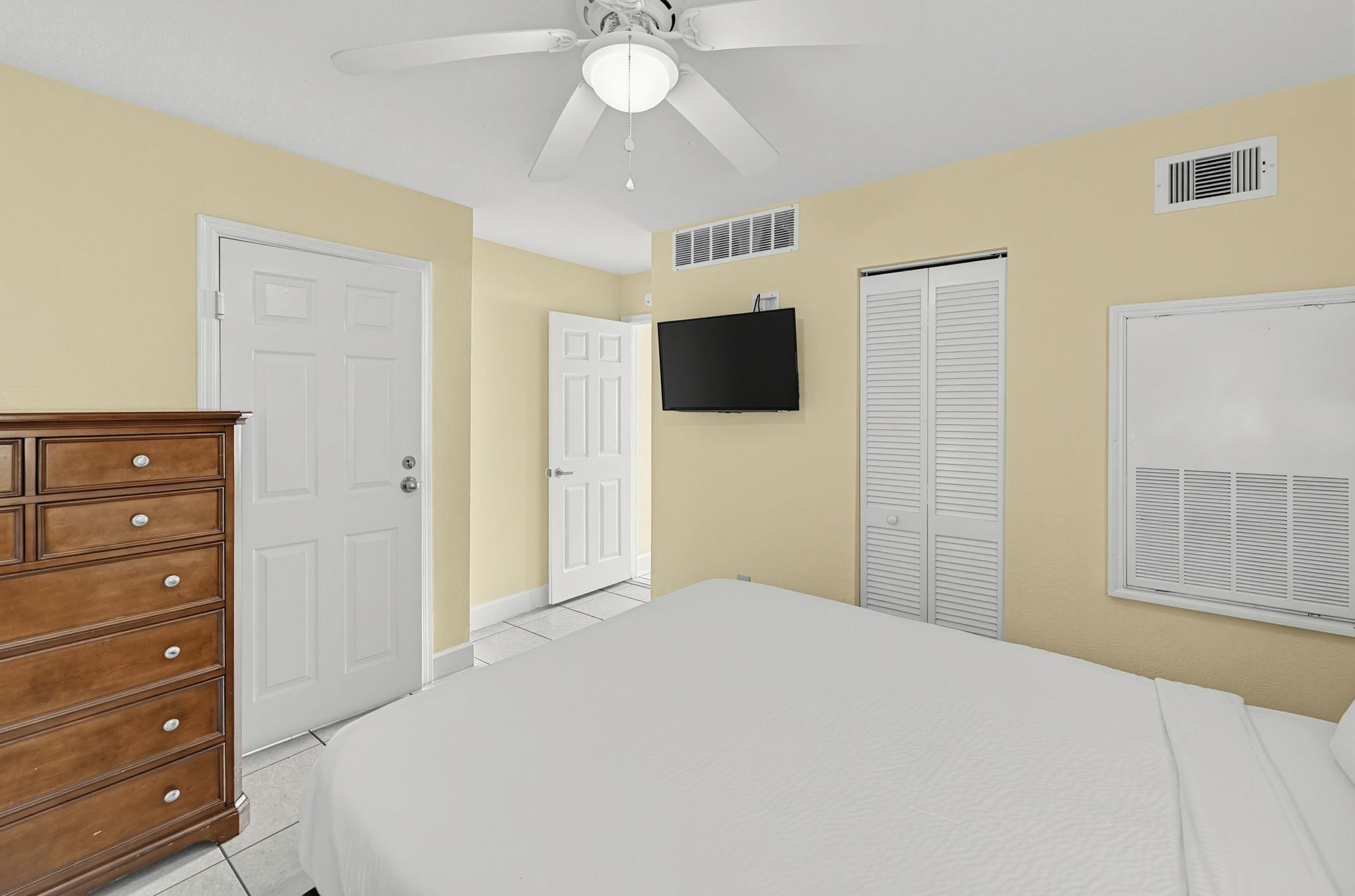 Quarto com cama, penteadeira, ventilador de teto, TV e duas portas.