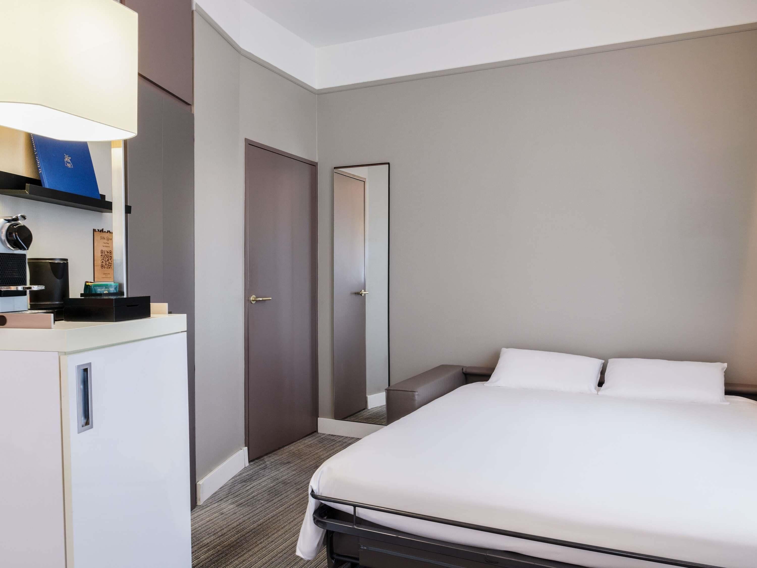 Quarto de hotel com cama, escrivaninha com luminária, cafeteira e porta.