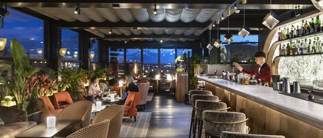 Uma área de bar moderna com balcão, assentos e vista da cidade ao entardecer.