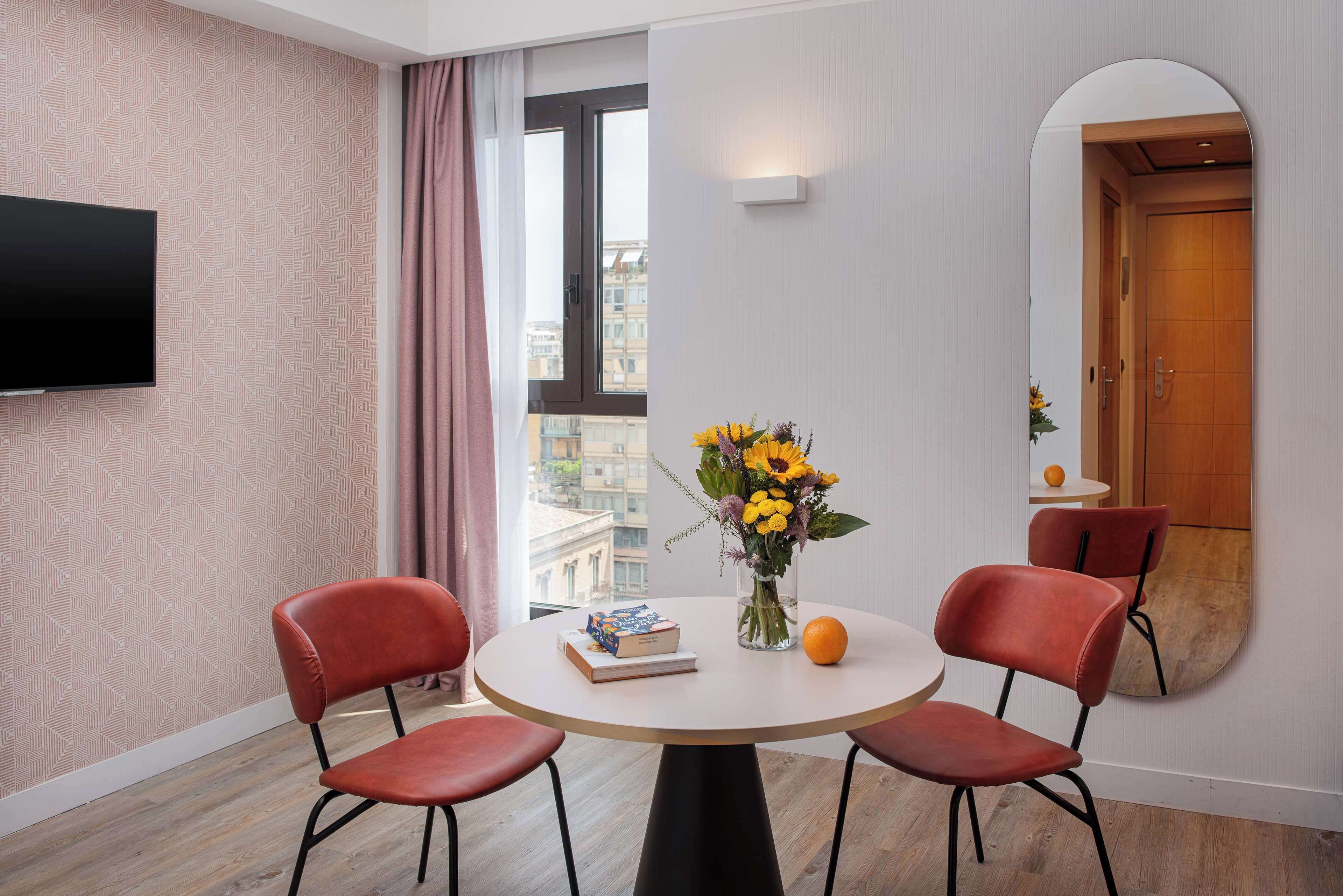 Quarto de hotel moderno com uma mesa redonda, duas cadeiras vermelhas, uma TV de tela plana, um vaso com flores e vista da cidade pela janela.