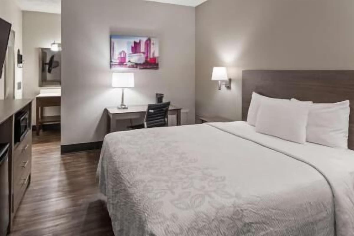 Quarto de hotel com cama, escrivaninha, cadeira, televisão e um quadro na parede.