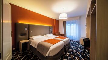 Quarto de hotel com duas camas, uma mesa de cabeceira, uma cadeira, uma escrivaninha e uma janela grande com cortinas.