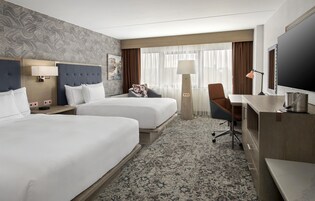 Quarto de hotel com duas camas, uma escrivaninha, uma cadeira, uma televisão e uma janela com cortinas.