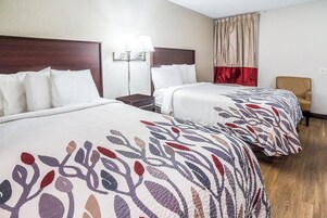 Quarto de hotel com duas camas, mesa de cabeceira, abajur, cadeira e janela com cortinas.