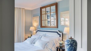 Uma cama bem arrumada com lençóis brancos e detalhes azuis, uma janela grande com vista para edifícios, e um vaso azul no criado-mudo.