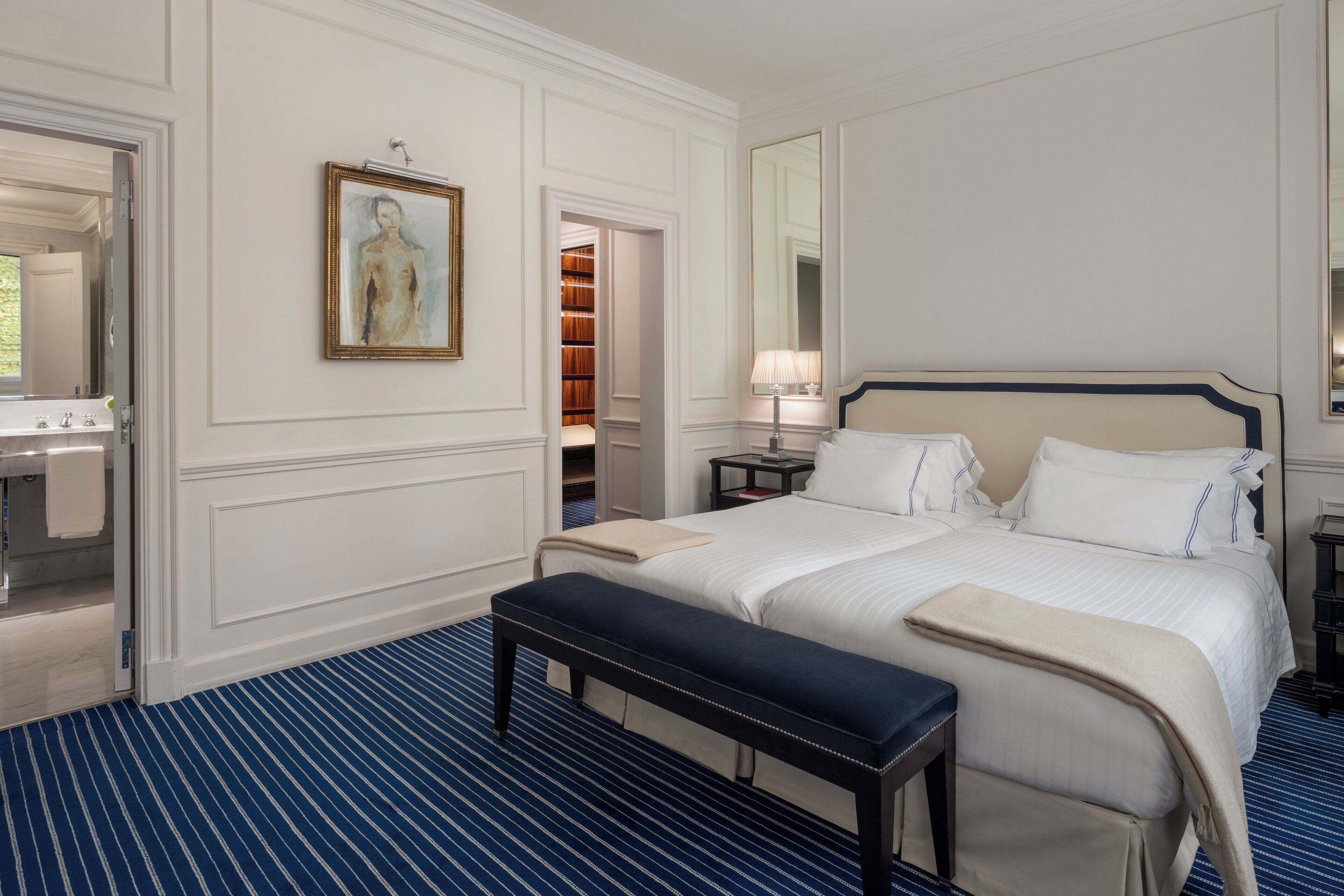 Quarto de hotel com uma cama grande, um banco, uma mesa de cabeceira, um espelho e um guarda-roupa.