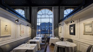 Um restaurante com várias mesas, vista para um edifício através de grandes janelas e obras de arte emolduradas nas paredes.