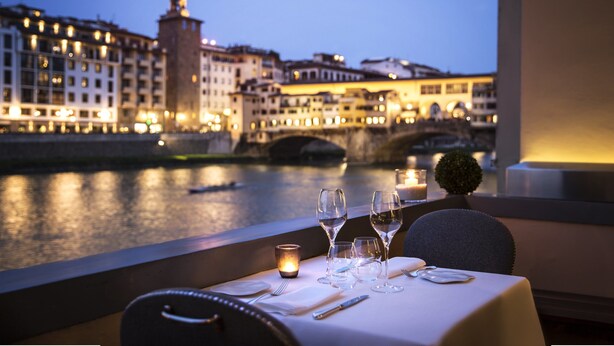Mesa posta para dois, com taças de vinho, vela e talheres, com vista para um rio e edifícios ao fundo, incluindo uma ponte.