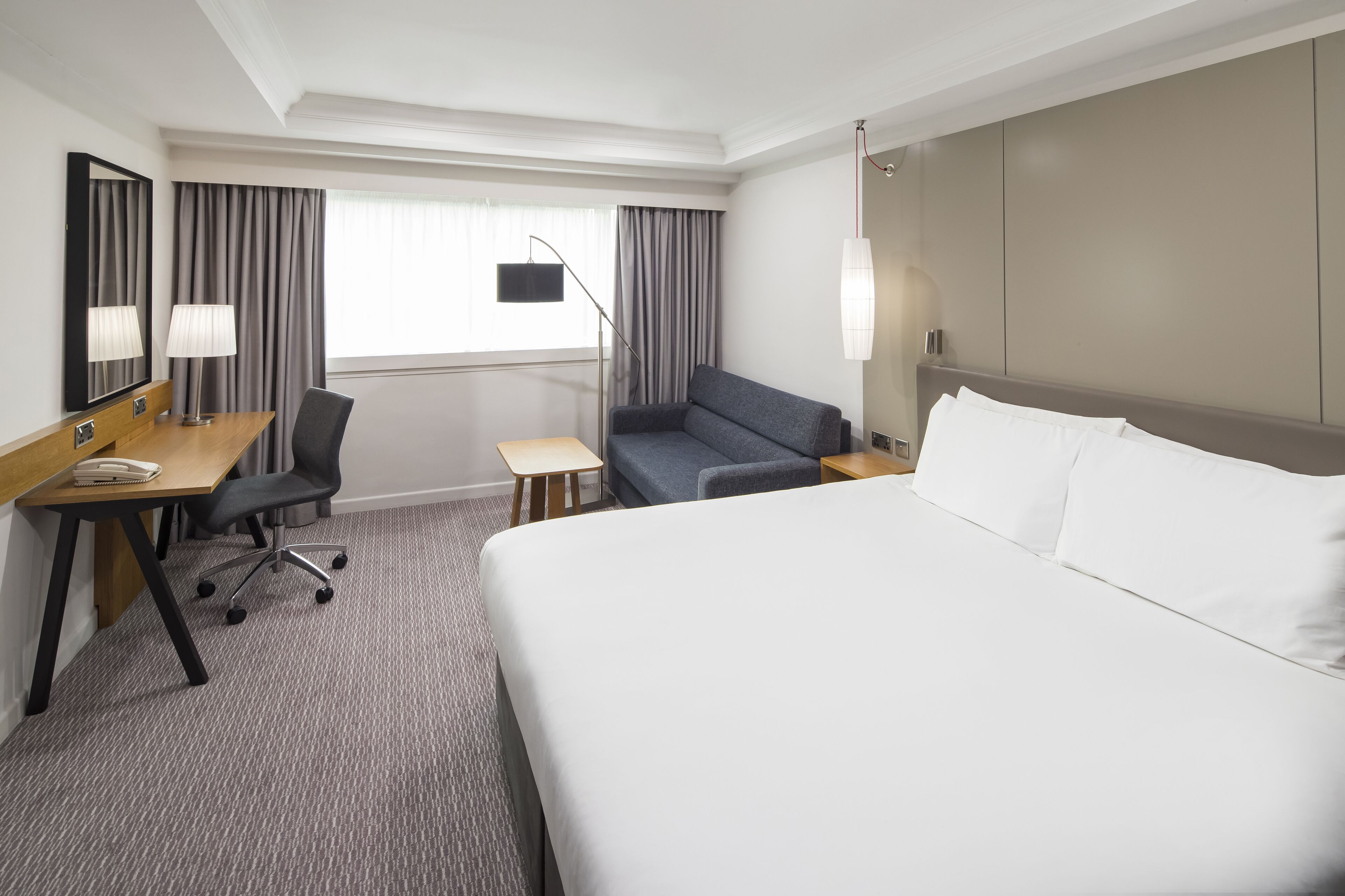 Quarto de hotel com uma cama grande, uma escrivaninha com cadeira, um sofá, uma mesinha e um espelho.