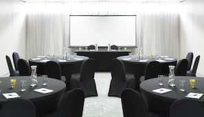 Uma sala de conferências com quatro mesas, cada uma com cadeiras, uma tela de projetor e garrafas de água.