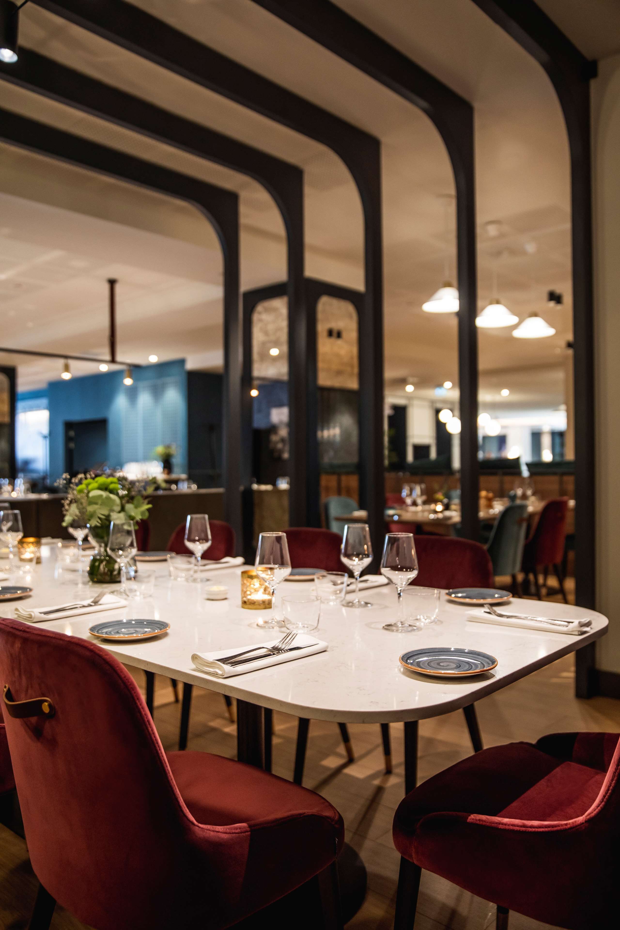 Uma sala de jantar com uma mesa branca posta para quatro pessoas, cadeiras de veludo vermelho e uma parede azul de destaque ao fundo.