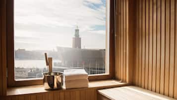 Uma sala de sauna com interior de madeira, uma janela com vista para a paisagem urbana, um balde de metal e uma toalha.