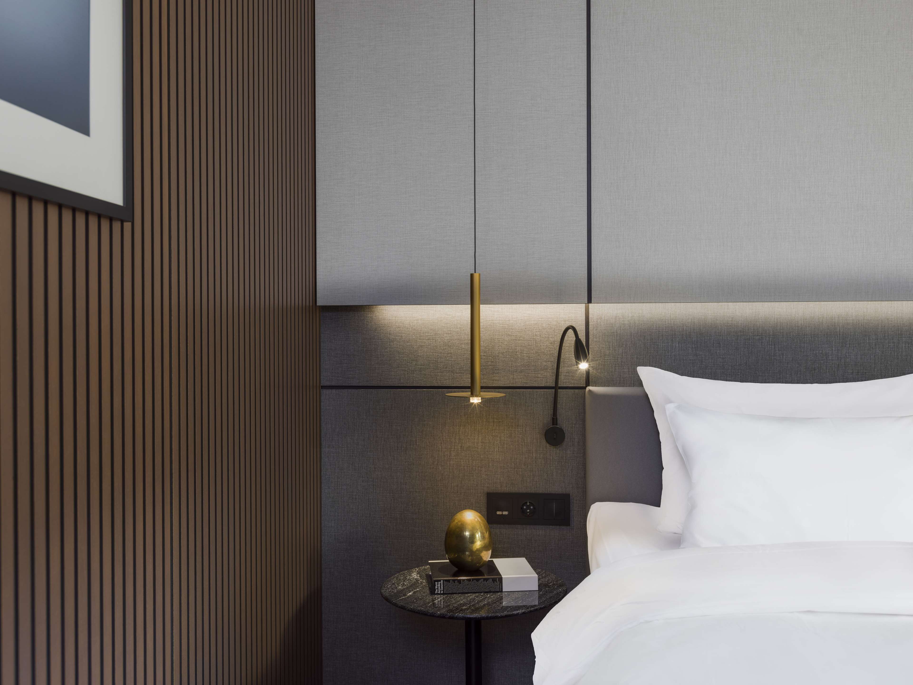 Quarto de hotel moderno com cama bem arrumada, mesa de cabeceira com abajur e painel de controle fixado na parede.