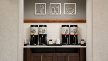 Uma estação de café com quatro dispensadores de café identificados como 'Light Roast', 'Dark Roast', 'Água Quente' e 'Descafeinado'. Acima da estação, há três obras de arte emolduradas retratando elementos relacionados ao café.