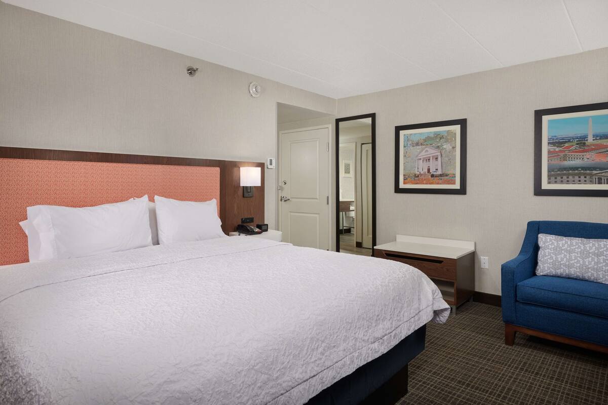 Quarto de hotel com uma cama grande, uma escrivaninha, uma cadeira e duas molduras na parede.
