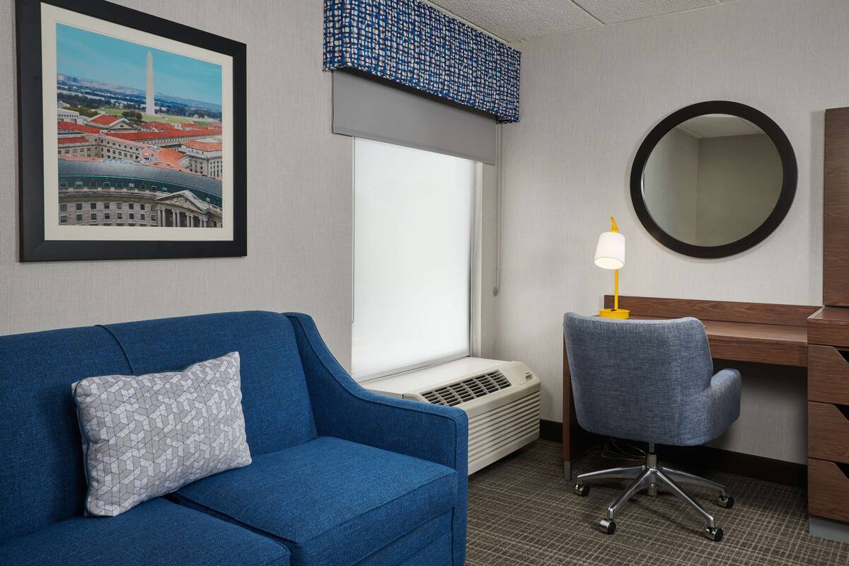 Quarto de hotel com sofá azul, uma escrivaninha com cadeira, uma janela grande com vista para edifícios, um quadro na parede e um espelho redondo.