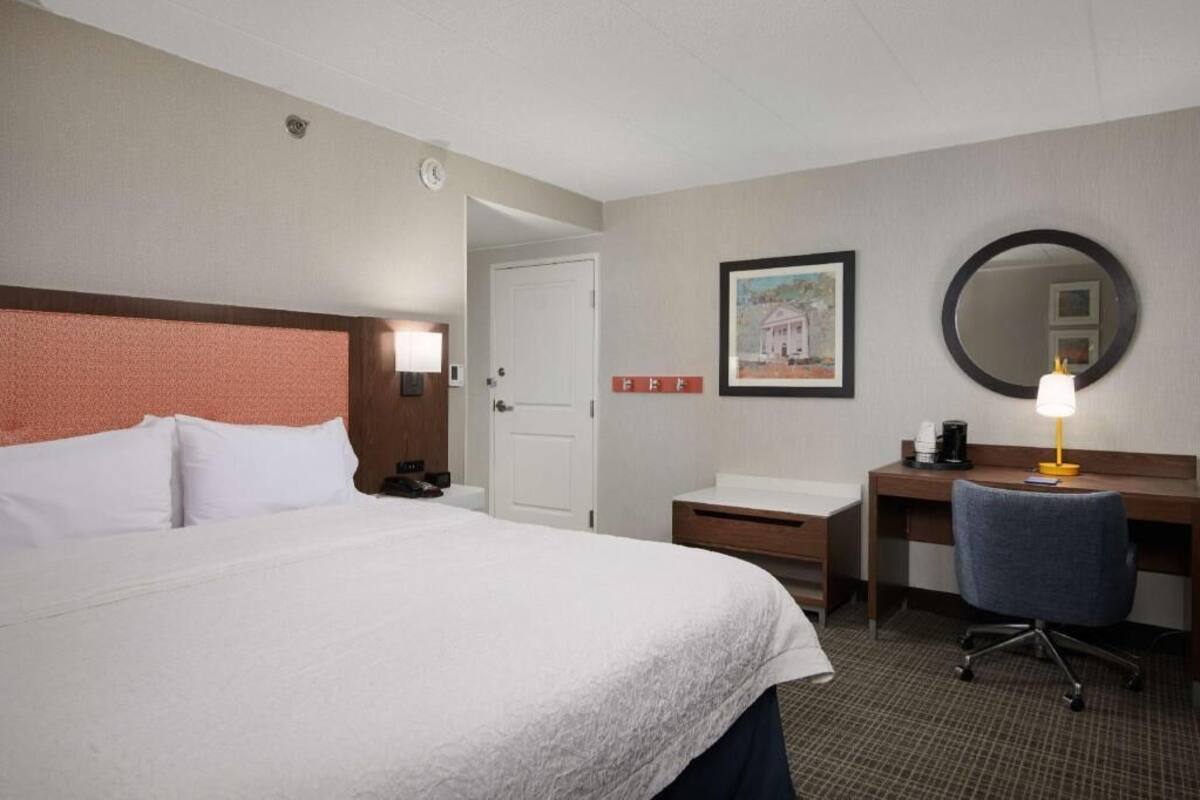 Quarto de hotel com uma cama, escrivaninha, cadeira, abajur, espelho e um quadro na parede.