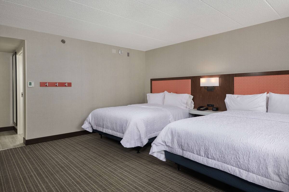 Quarto de hotel com duas camas, piso coberto por carpete, cabeceira de madeira, mesa de cabeceira e luminária fixada na parede.