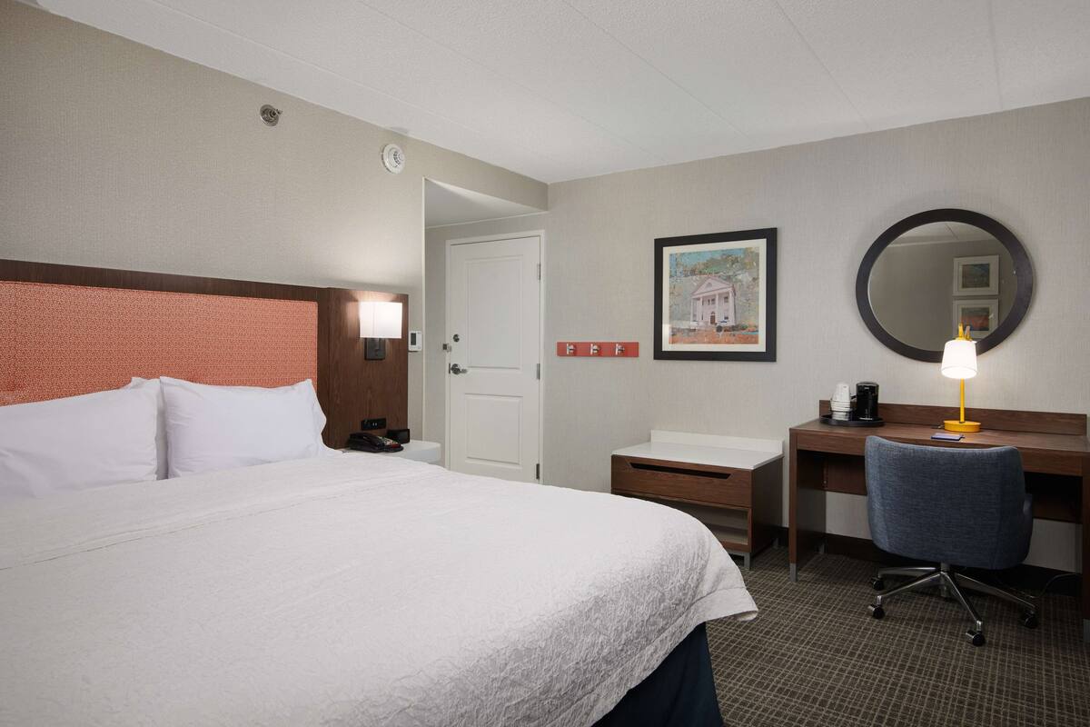 Quarto de hotel com uma cama grande, uma escrivaninha, uma cadeira, abajur, espelho e uma porta.