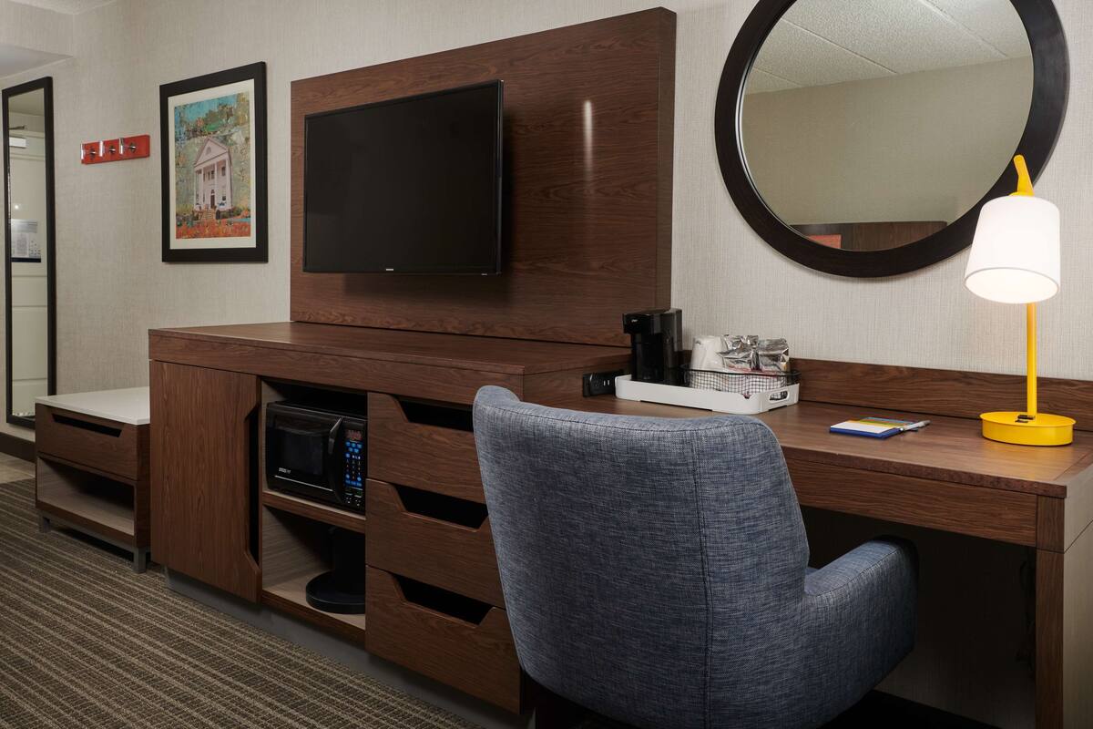 Quarto de hotel com escrivaninha de madeira, poltrona estofada cinza, televisão de tela plana, micro-ondas, cafeteira e abajur.