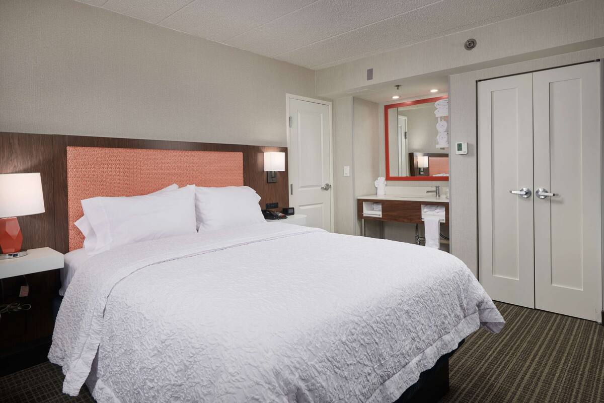 Quarto de hotel com uma cama grande, uma mesinha de cabeceira com luminária, banheiro com espelho e um guarda-roupa.