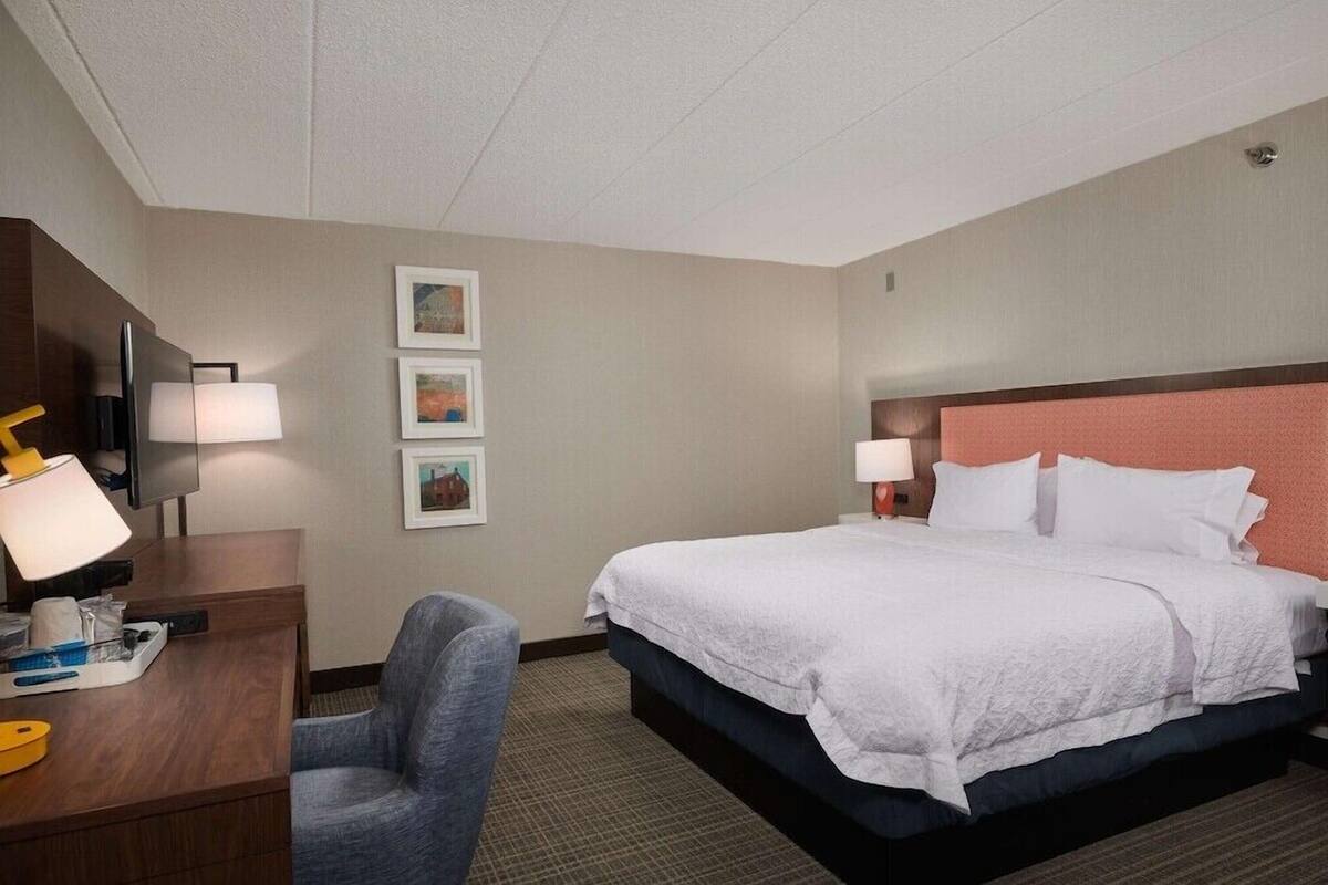 Quarto de hotel com cama, escrivaninha com luminária, cadeira e quadros nas paredes.
