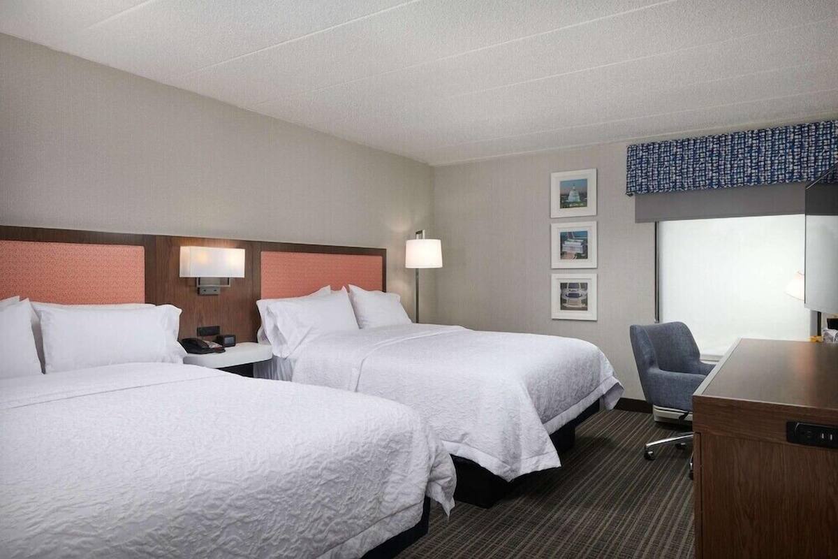 Quarto de hotel com duas camas, uma escrivaninha, uma cadeira e uma janela com vista.