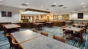 Uma área de refeições de hotel com várias mesas e cadeiras, uma receção e vista para a cidade através de grandes janelas.