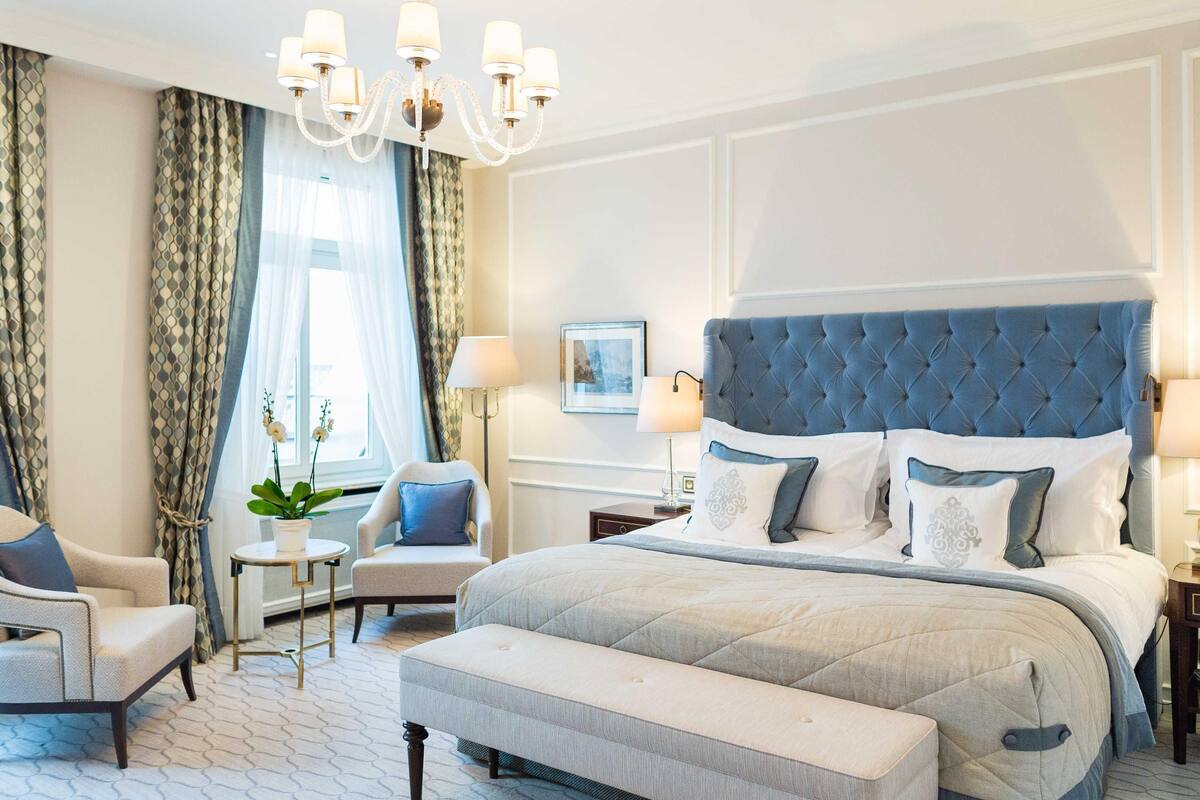 Quarto de hotel com uma cama grande, área de estar com uma poltrona e uma mesa pequena, um candelabro e cortinas.
