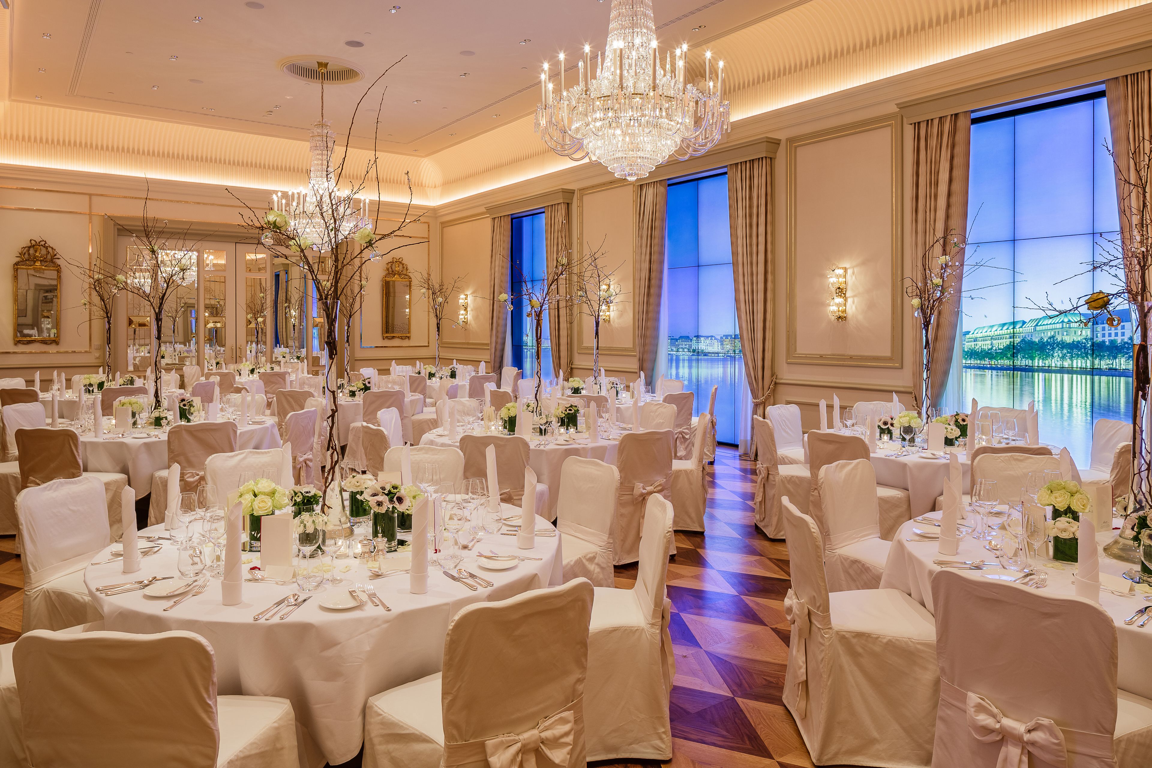 Uma sala de banquetes com mesas redondas, toalhas brancas e cadeiras, decorada com arranjos florais e velas.