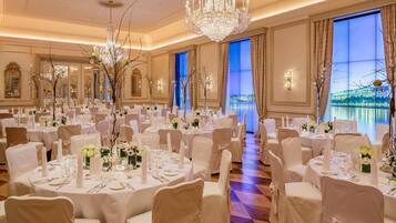 Uma sala de banquetes com mesas redondas, toalhas brancas e cadeiras, decorada com arranjos florais e velas.