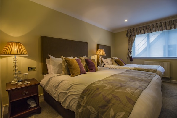 Quarto de hotel com duas camas, uma mesa de cabeceira, luminárias e uma janela com cortinas.