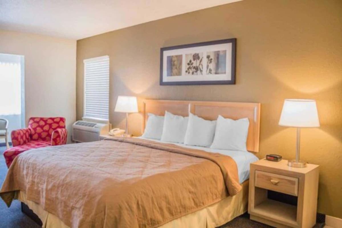 Quarto de hotel com cama, mesas de cabeceira, uma cadeira, janela com persianas e um quadro na parede.