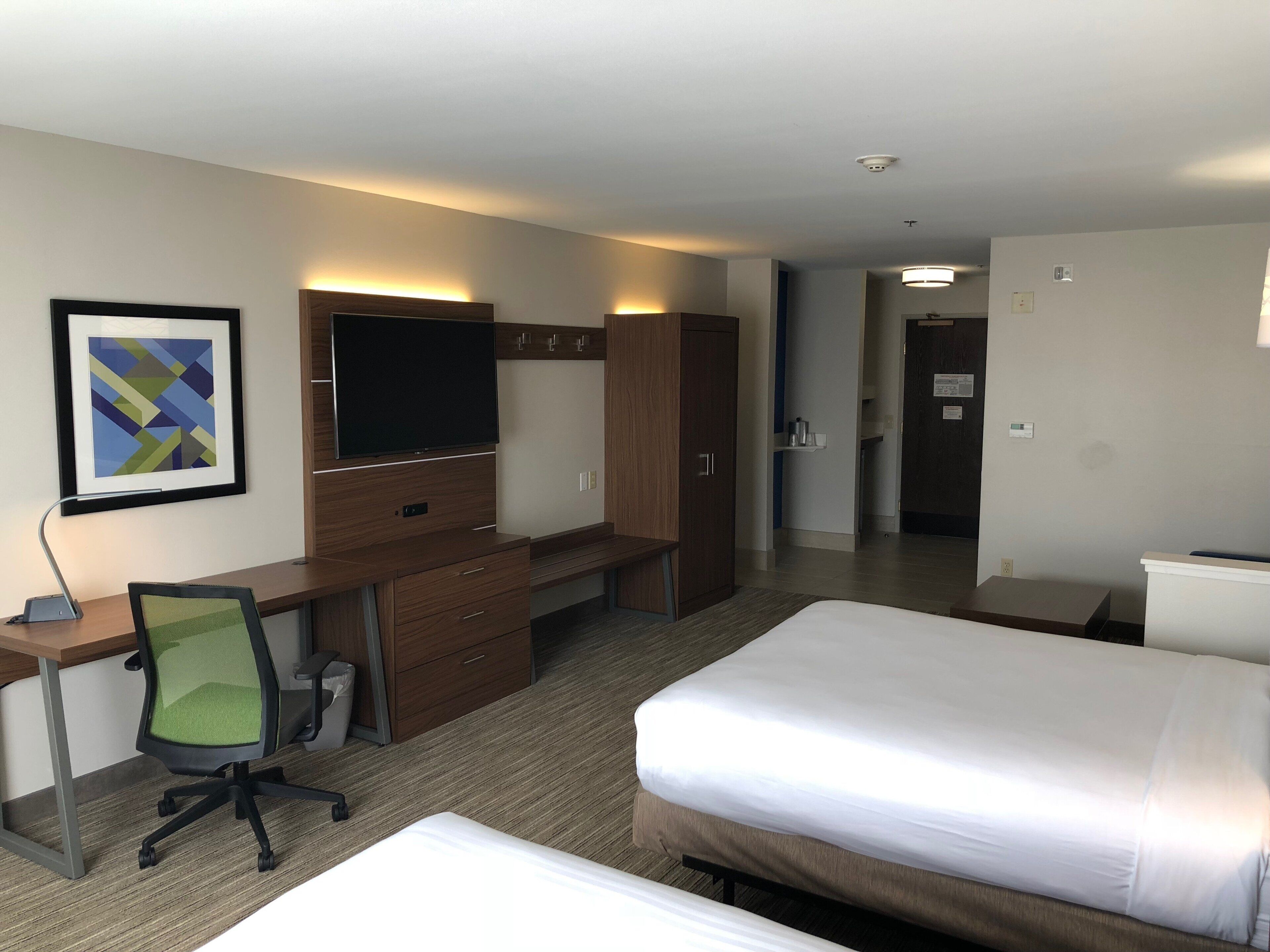 Quarto de hotel com cama, escrivaninha com cadeira, televisão e guarda-roupa.