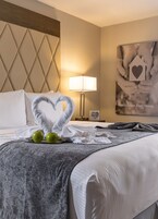 Uma cama bem arrumada com toalha em formato de coração e maçãs, uma luminária de cabeceira e um quadro na parede.