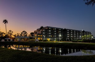 Um hotel com vários andares, piscina na frente, cercado por vegetação e palmeiras.