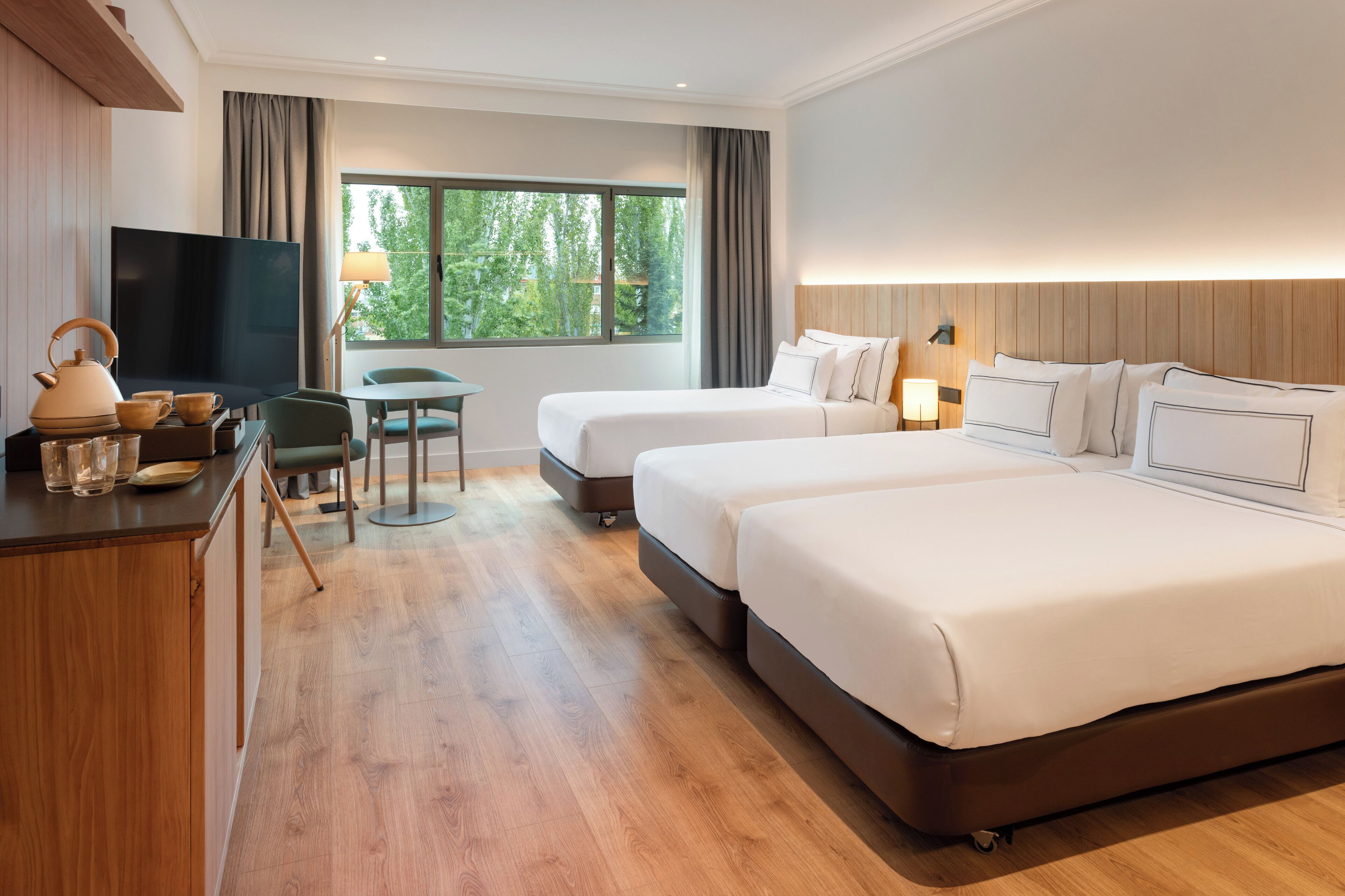 Quarto de hotel com duas camas, televisão, secretária com chaleira e janela com vista para árvores.