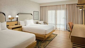 Quarto de hotel com duas camas, uma mesa, uma cadeira, uma janela grande com cortinas e uma luminária fixada na parede.